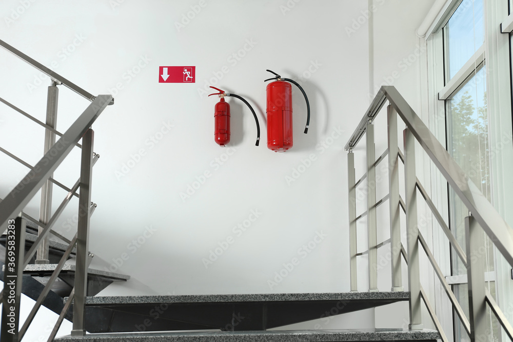 Fire extinguishers and emergency exit sign on white wall near staircase ...