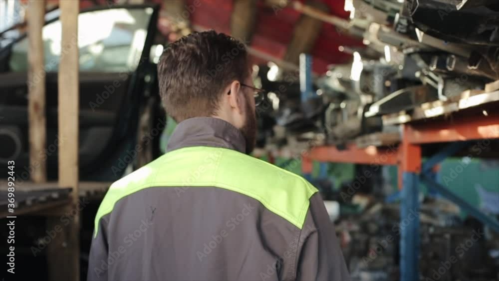 A young auto mechanic in overalls walks between a rack of spare parts ...