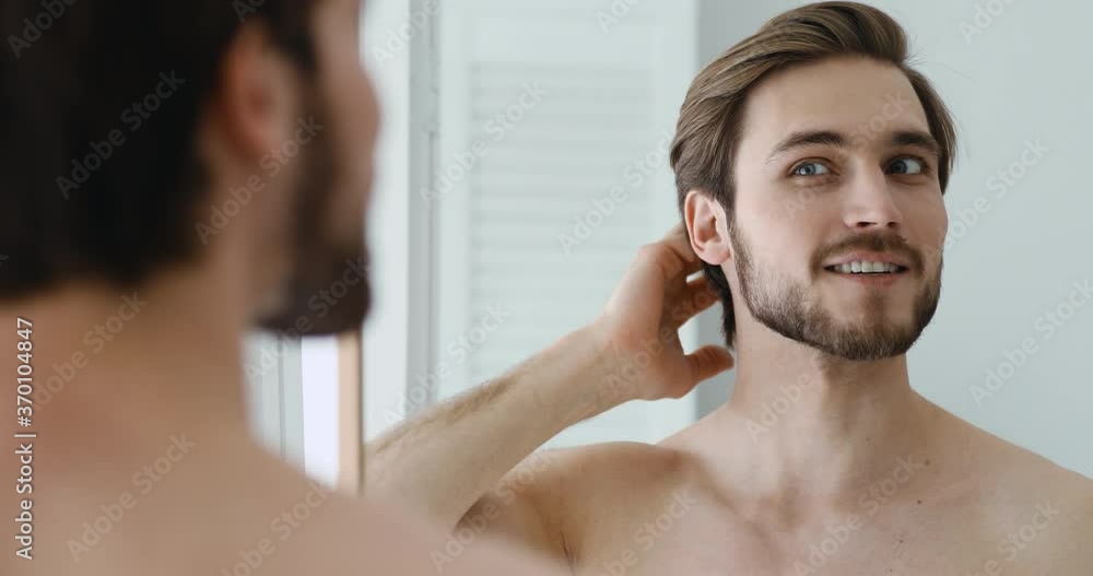 Smiling shirtless caucasian man looking at mirror, combing hair with ...