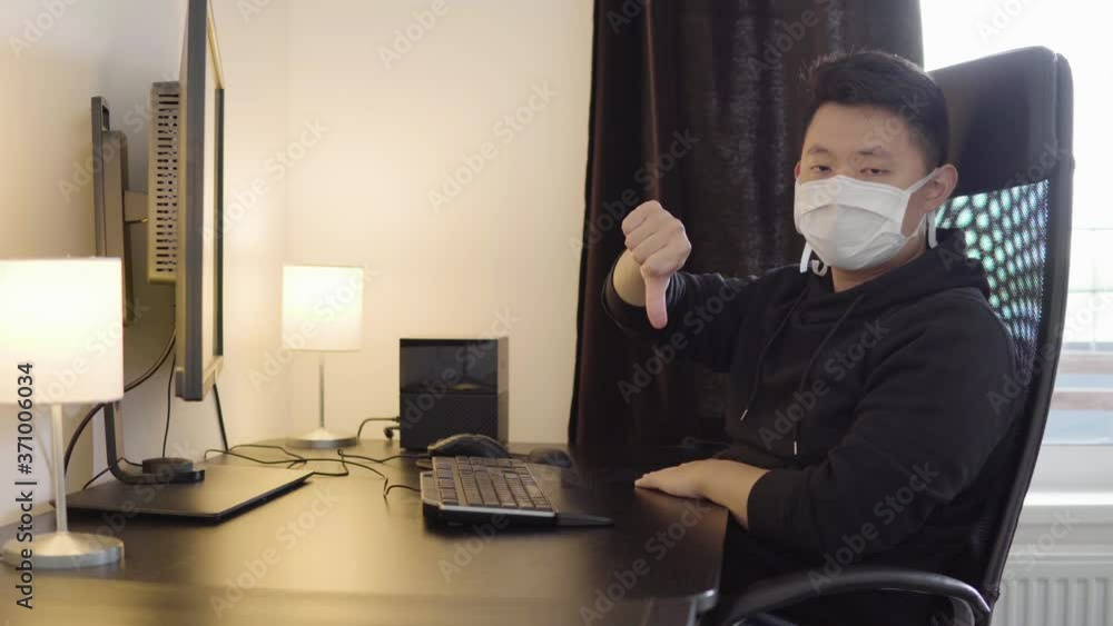 A young Asian man in a face mask looks at the desktop computer screen ...