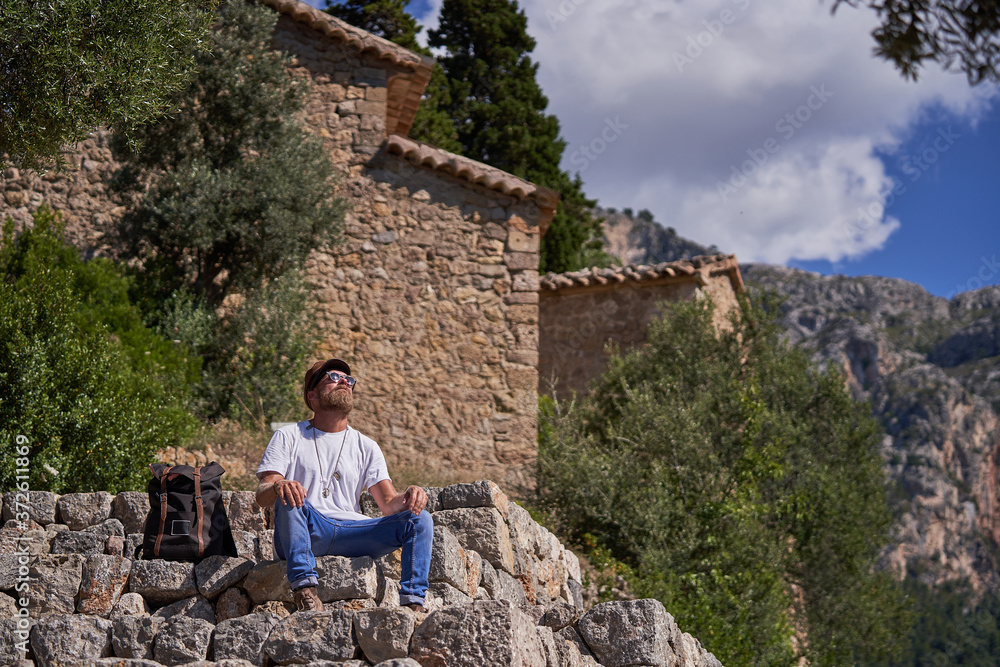 Contemplative hipster male backpacker sitting on stone border near ...