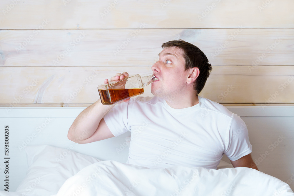 Drunk rowdy man with angry face, bottle of alcohol sits drinking in bed ...