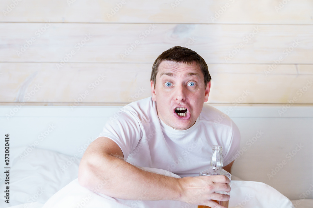 Drunk rowdy man with angry face, bottle of alcohol sits drinking in bed ...