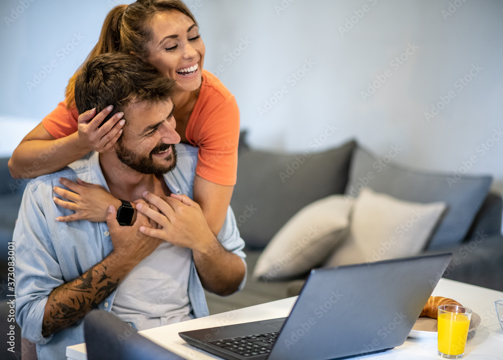 A woman hugs her husband who works on a laptop, they both smile Stock ...