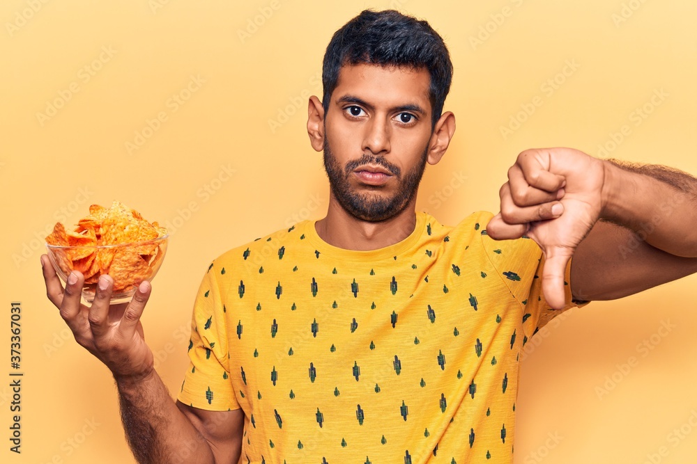 Young latin man holding nachos potato chips with angry face, negative ...