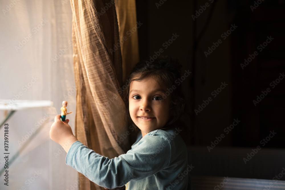 Little girl plays with a doll next to the window of her living room ...