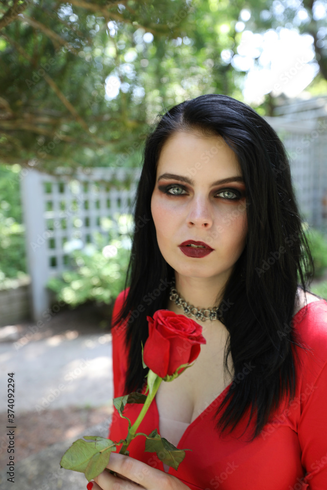 Gothic woman in red dress sitting in a garden with a rose. Beautiful young woman with bright ...