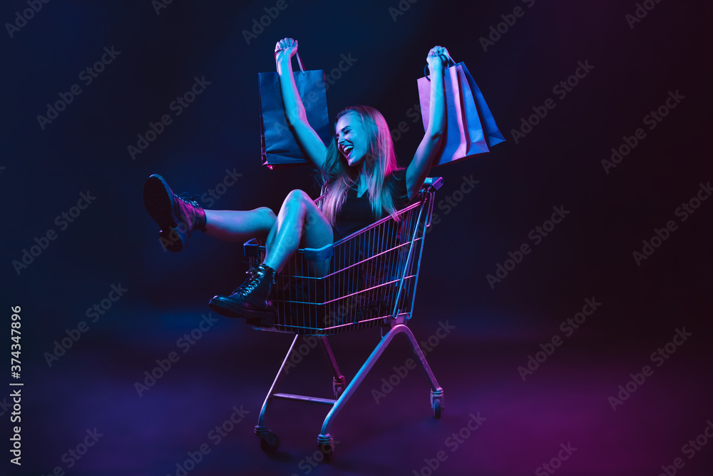 Rolling in a cart with shopping bags. Portrait of young woman in neon ...