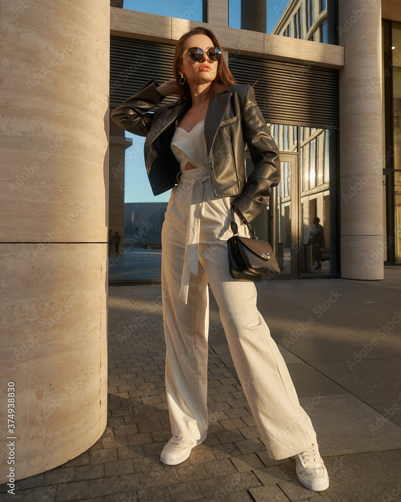 Young stylish woman in white overall, sneakers, black leather jacket ...