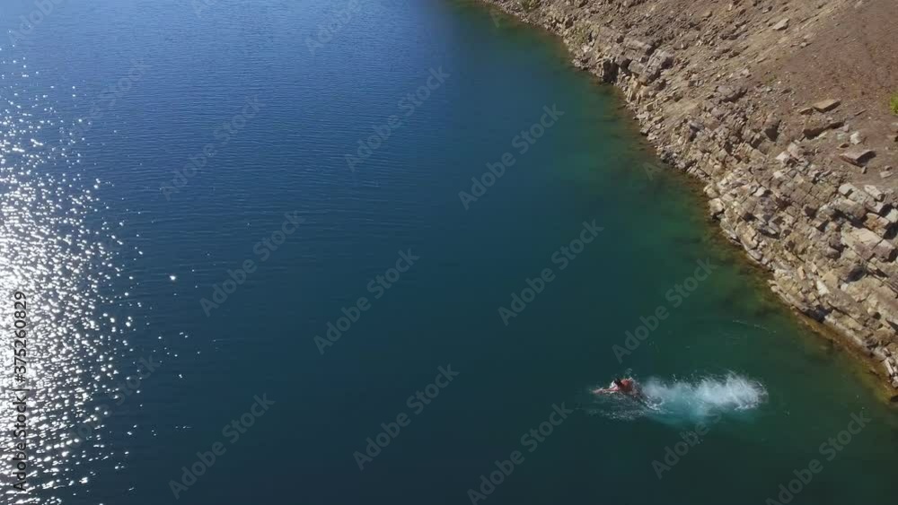 Aerial view. A muscular man jumps off a cliff into the crystal clear ...