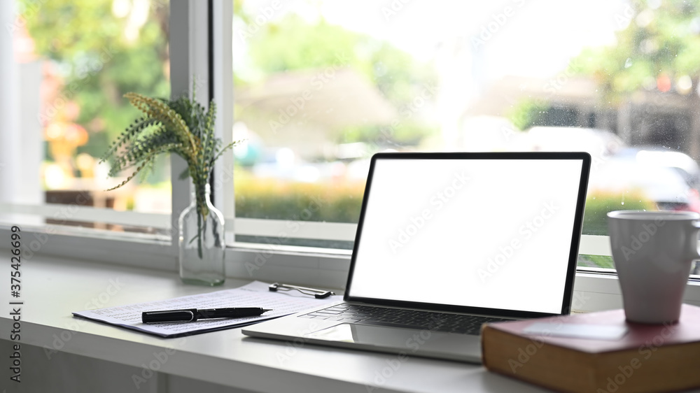 A computer laptop with a white blank screen is putting on a windowsill ...