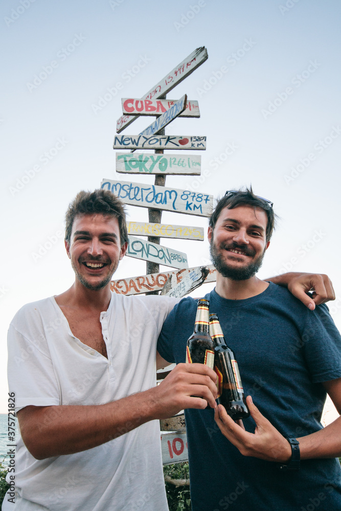 Two young men friends toasting beers next to a signpost with directions ...