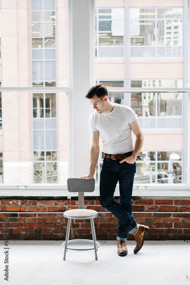 young male standing next to chair indoors by large windows and white ...