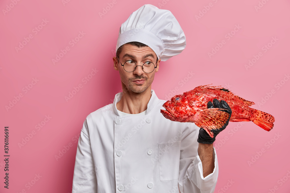 Handsome skilled male cook looks attentively at big fish, thinks what ...