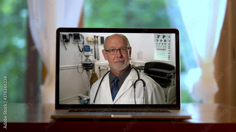 Laptop set on desk in front of french doors with a male doctor talking ...