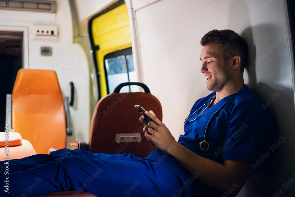 Young doctor sitting with his smartphone in the ambulance car at night ...