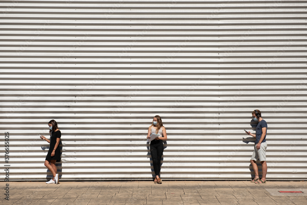 Three young students waiting in line on a queue wearing face masks and ...