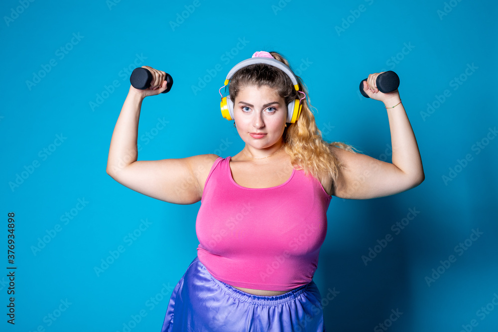 Plus size young black woman weight lifting on blue background listening ...