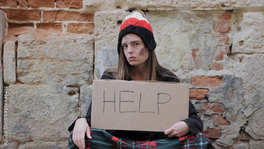 A homeless girl in a hat sits on the sidewalk and asks for help ...