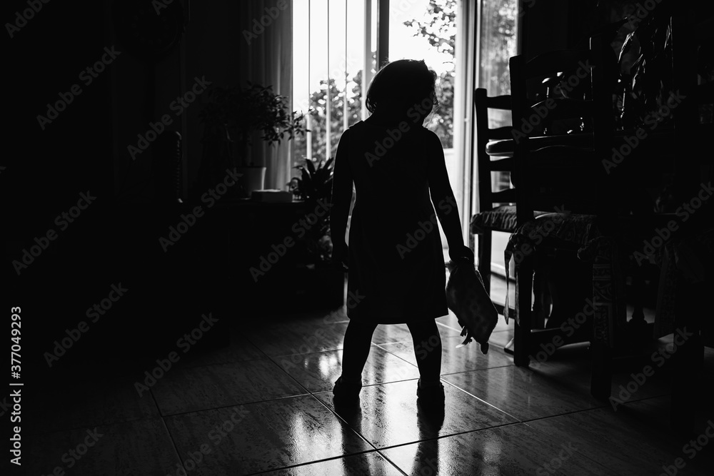 Little girl against the light with her rag doll in her hand in a scary ...