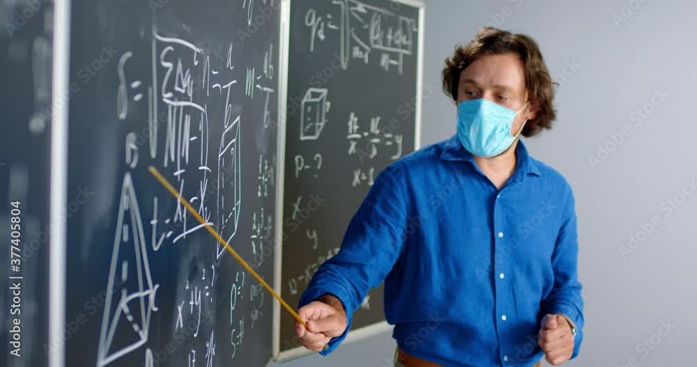 Caucasian male teacher in medical mask standing at board in classroom and telling physics or ...
