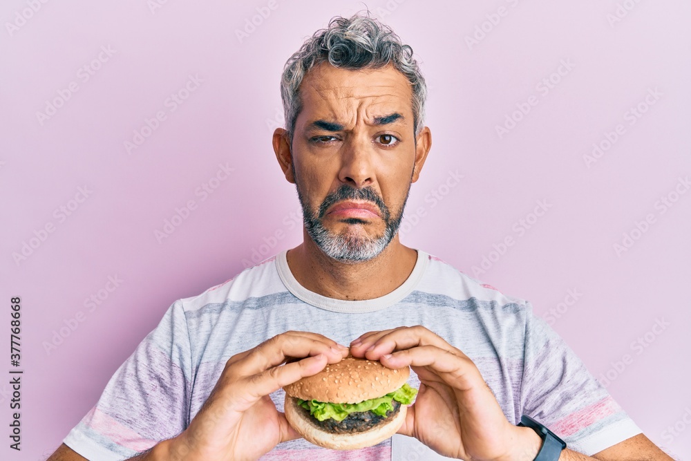 Middle age grey-haired man eating a tasty classic burger depressed and ...