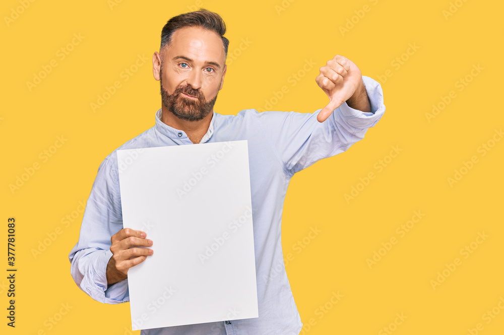 Handsome middle age man holding blank empty banner with angry face ...