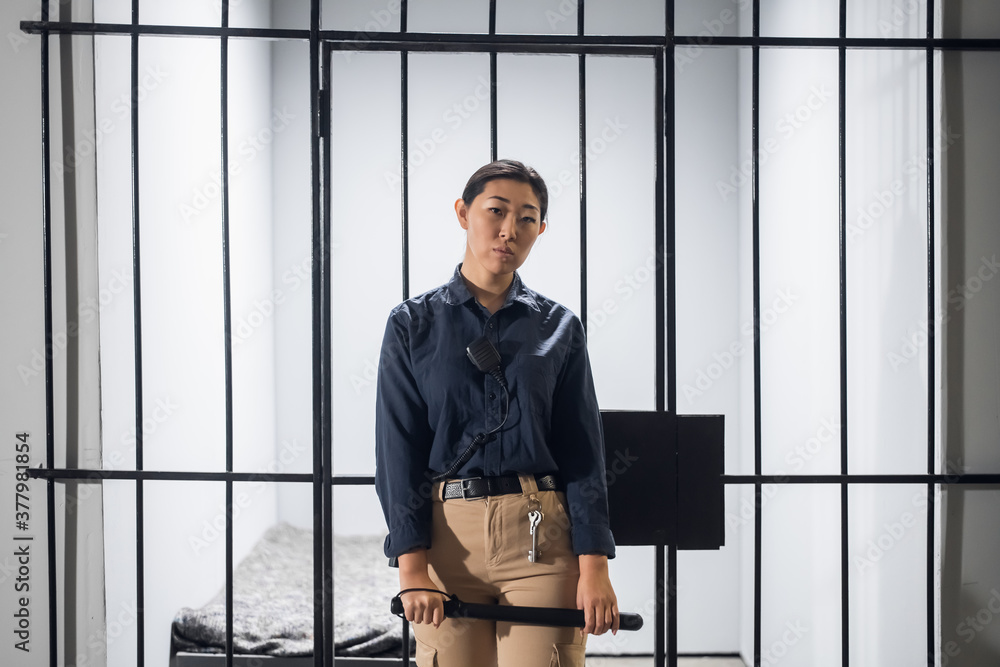 A young prison guard poses in front of prison bars in uniform. In a ...
