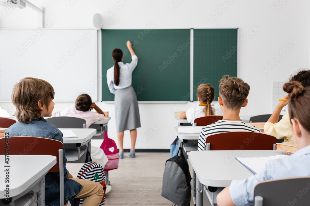 back view of teacher writing on chalkboard near multicultural pupils in ...