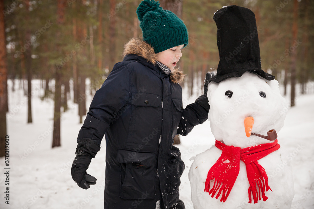 Cute boy builds a big snowman in the forest. Snowman is dressed in a ...