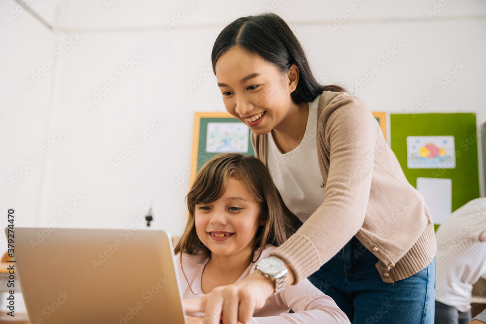 Young Asian teacher watching Caucasian school girl using computer in ...