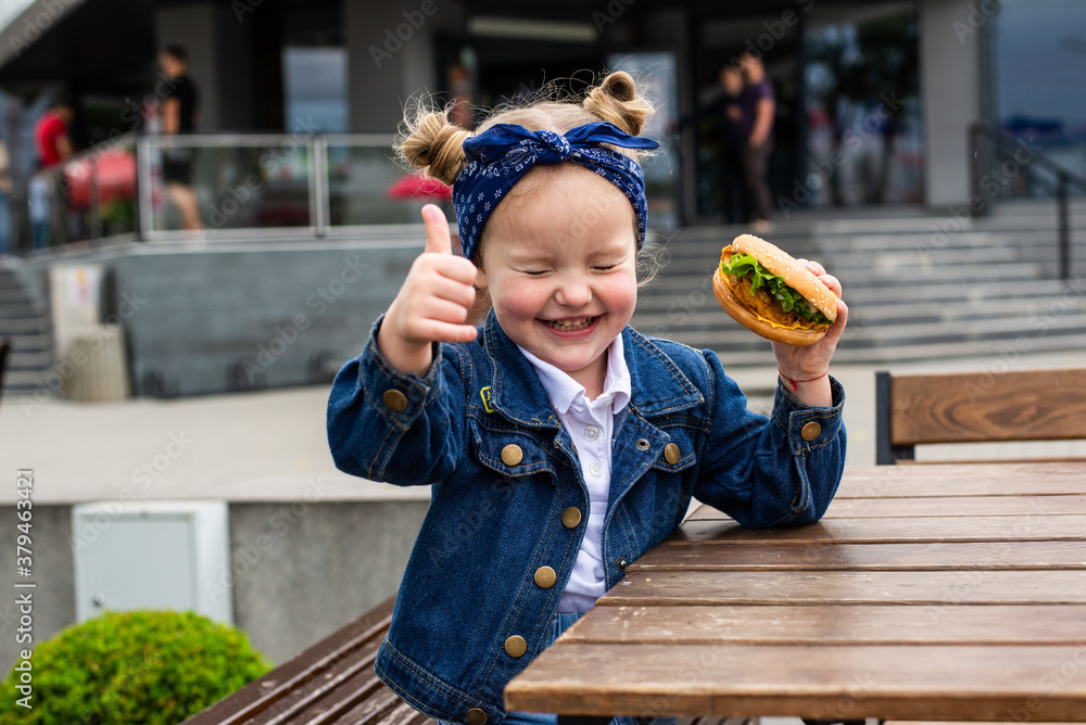 Little cute girl eating a burger showing thumbs up in a cafe. Concept ...