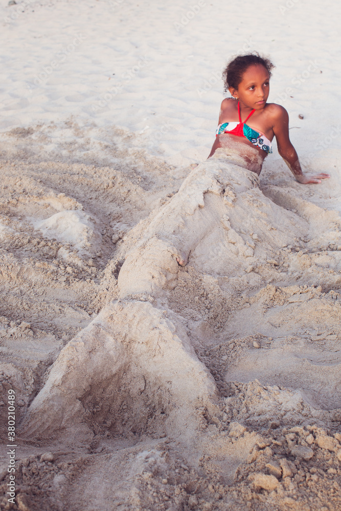 Young girl sitting on a beach with a mermaid tail made out of sand ...