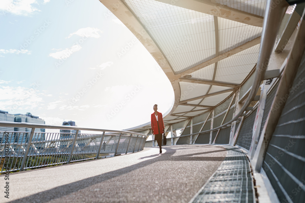 Elegant black woman in red jacket walking on geometric bridge ...