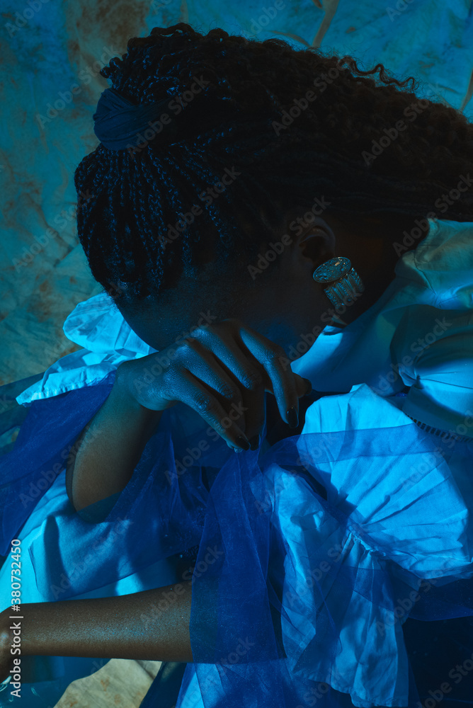 Sad black girl with braids and sleeves bows her head in a blue light ...