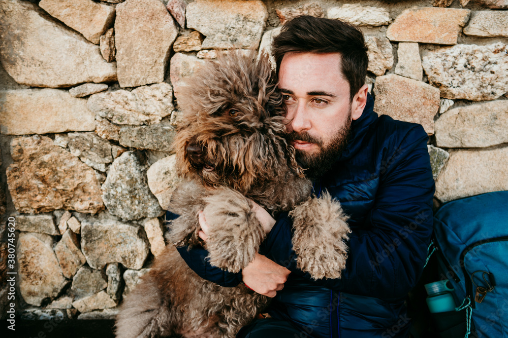 Bearded man embracing furry Labradoodle while sitting near stone wall ...