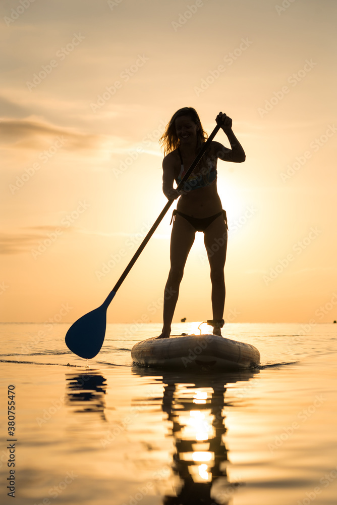 Back view of silhouette of unrecognizable female surfer standing on paddleboard and rowing ...
