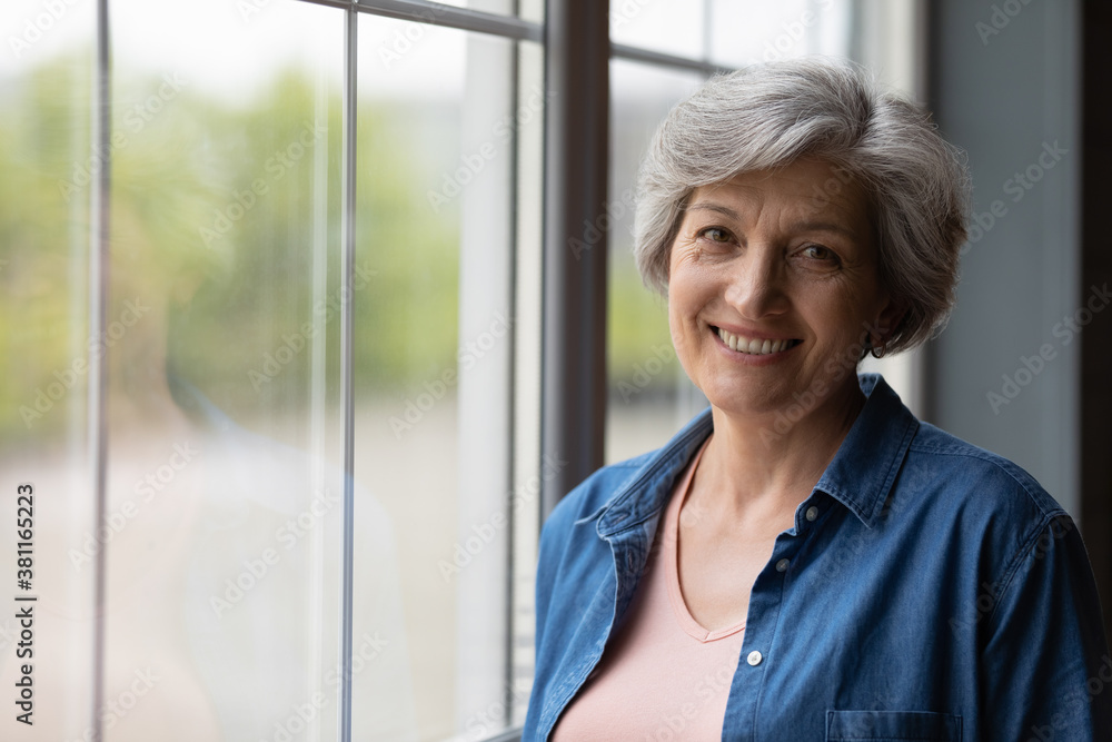 Portrait of smiling mature Caucasian 60s woman stand near window in own ...