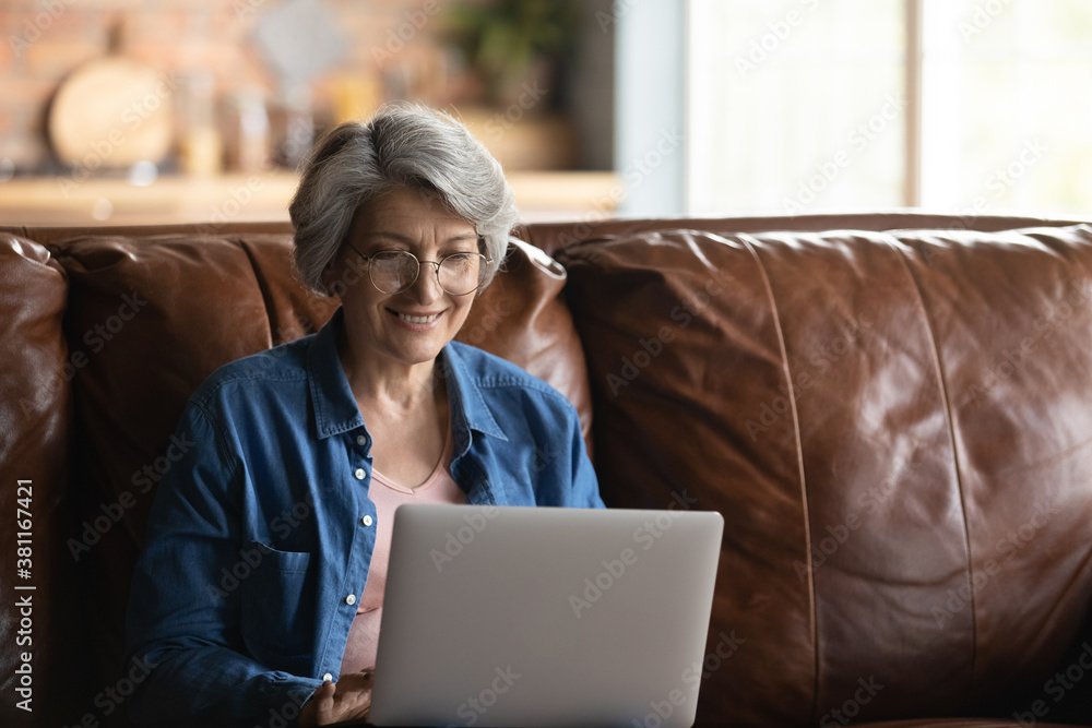 Happy modern senior 60s grandmother in glasses using laptop browsing ...