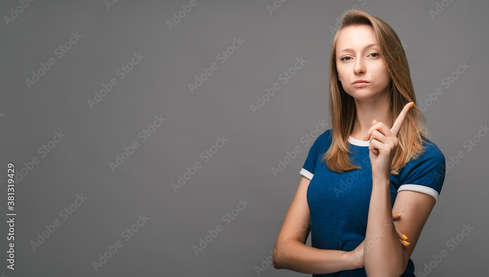Serious girl raises finger up says: Attention please. Studio shot gray ...