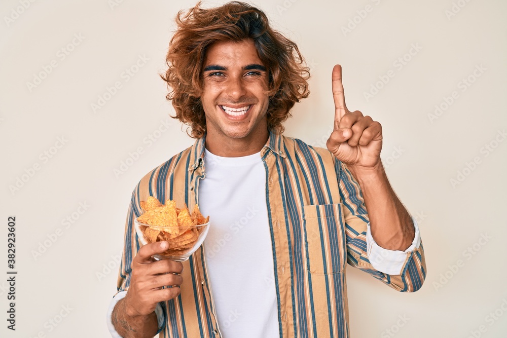 Young hispanic man holding nachos potato chips smiling with an idea or ...