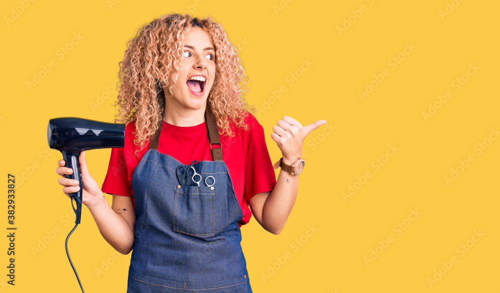 Young blonde woman with curly hair wearing hairdresser apron and holding dryer blow pointing ...