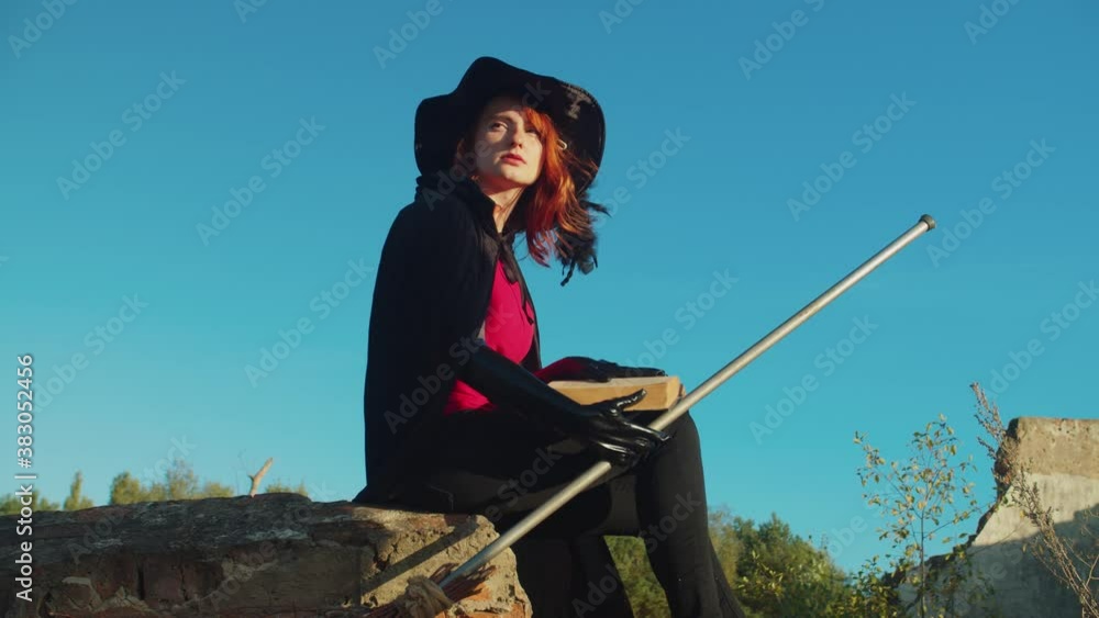 Evil attractive redhead witch in black cap and cloak with broom sitting ...