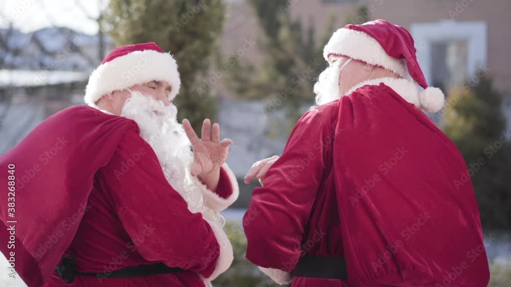 Two Santas in Christmas costumes with gift sacks turning, waving at ...