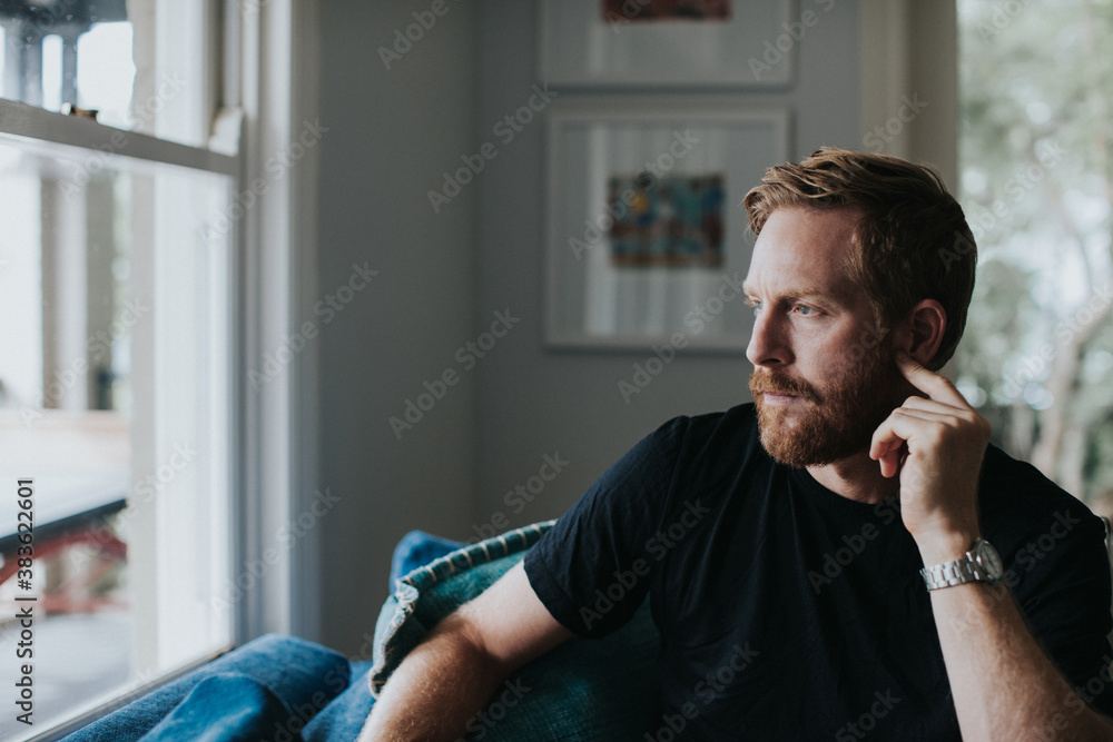 Thoughtful man looking through window while sitting on sofa at home ...