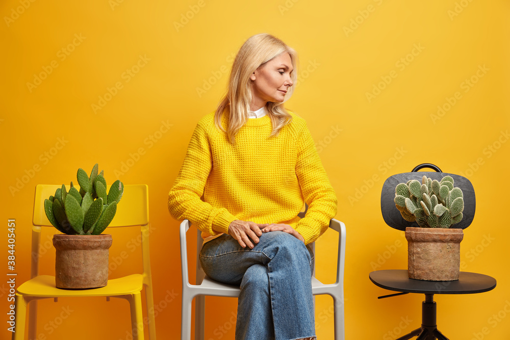 Calm senior woman poses between two chairs with cactus likes house plants isolated over yellow ...