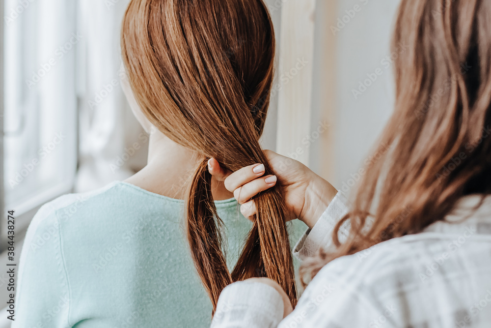 Two girls braid their hair at the window. Woman makes a braid to her ...