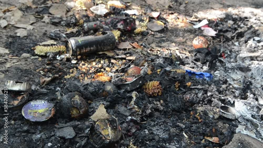 Broken glass bottles, food remains, cans and garbage in burned trash. A ...
