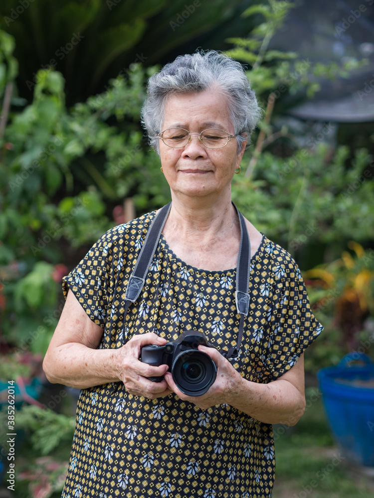 Elderly woman holding digital camera while standing in a garden. Asian ...