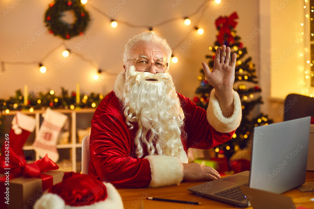 Santa Claus looking at camera and waving hand while working on laptop ...